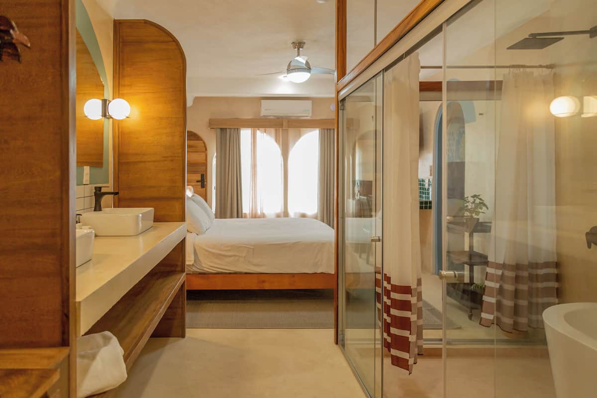 Quarto de hotel moderno com cama, box de vidro para o chuveiro, toucador com espelho e ventoinha no teto.