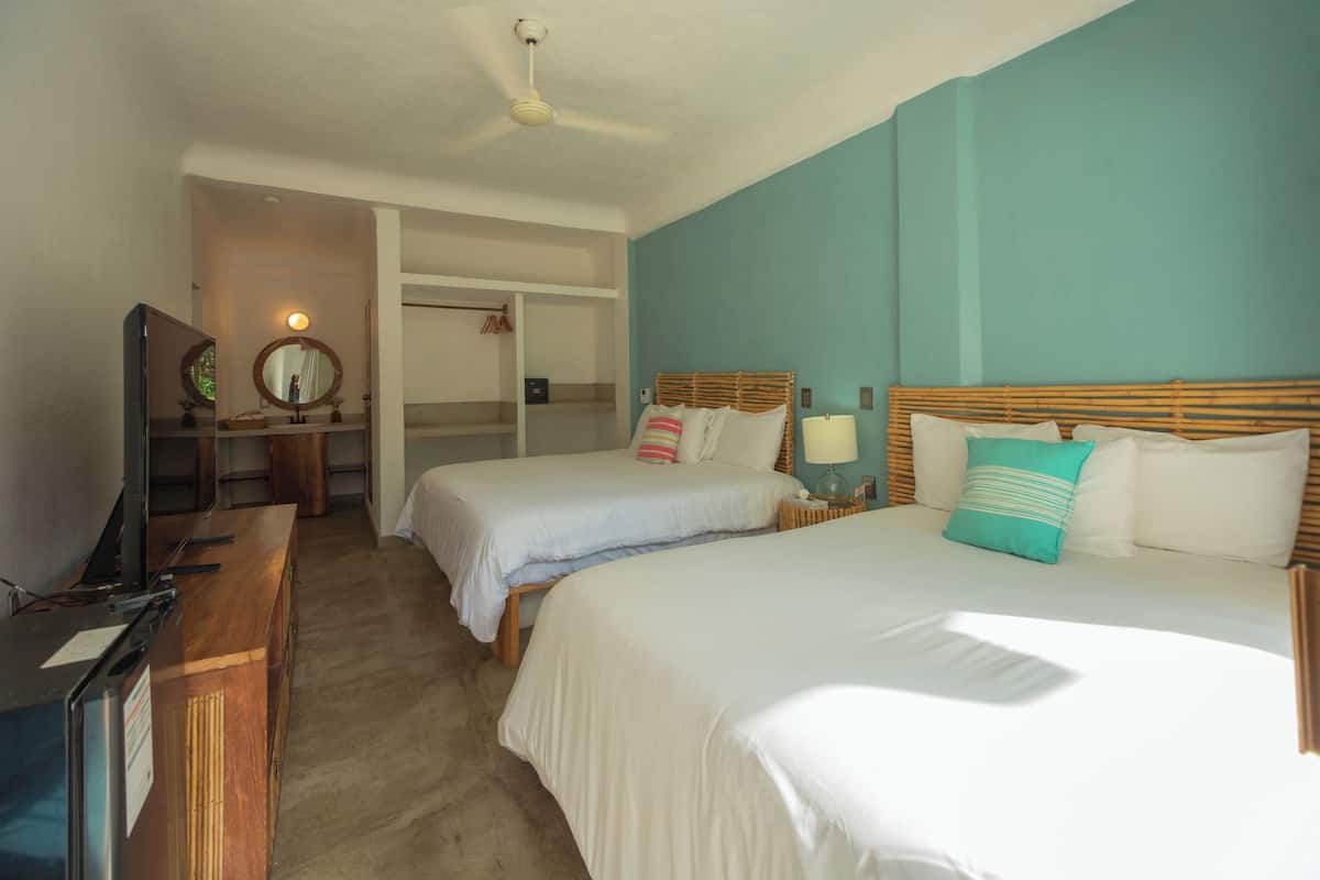 Quarto de hotel com duas camas, uma escrivaninha, um espelho e um ventilador de teto.