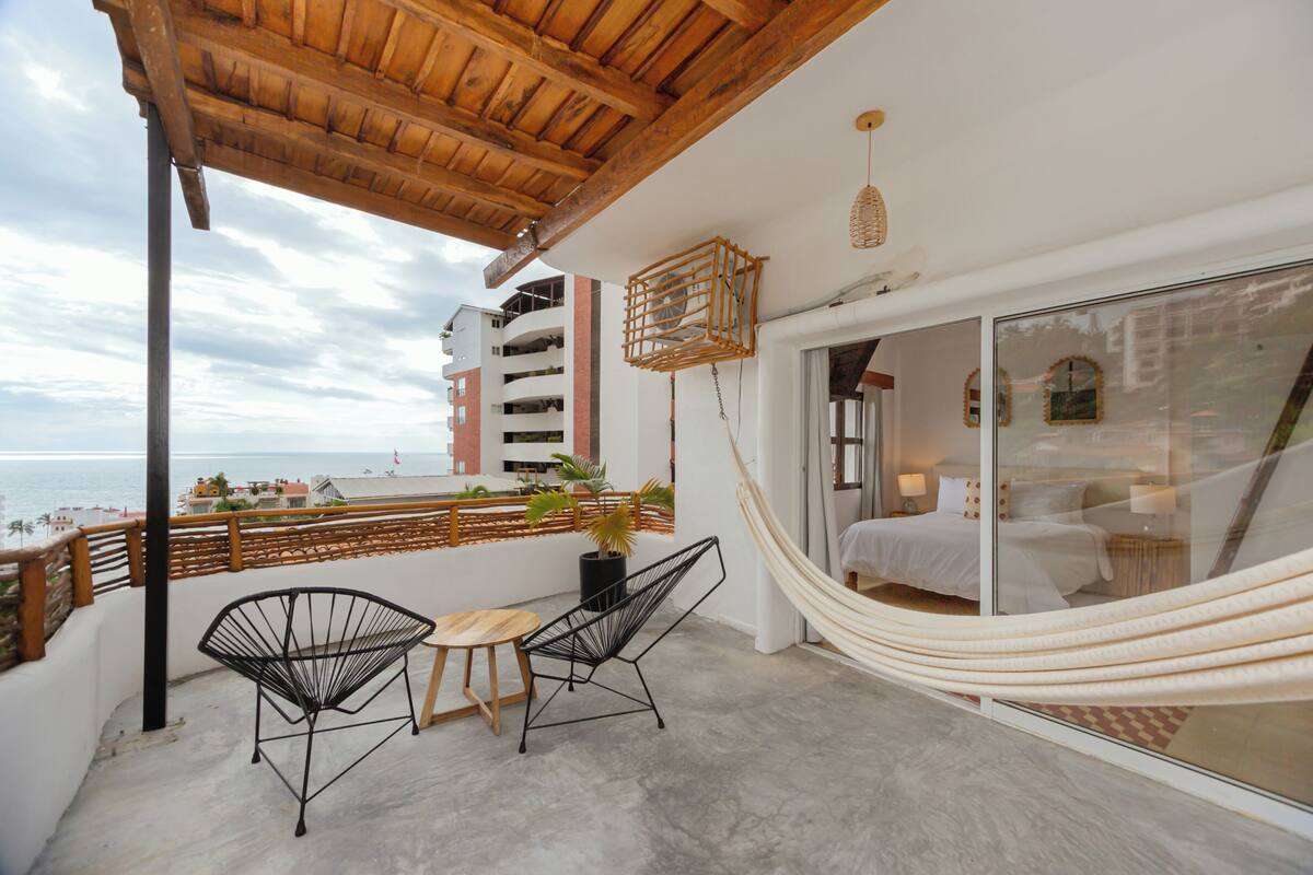 Uma varanda com teto de madeira, uma rede, duas cadeiras, uma mesa e vista para o mar.