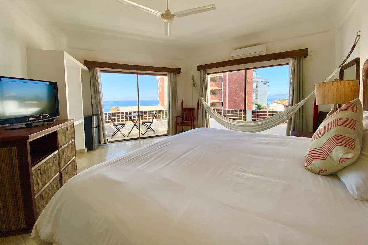 Quarto de hotel com cama, televisão, ventilador de teto, rede e varanda com vista para o mar.