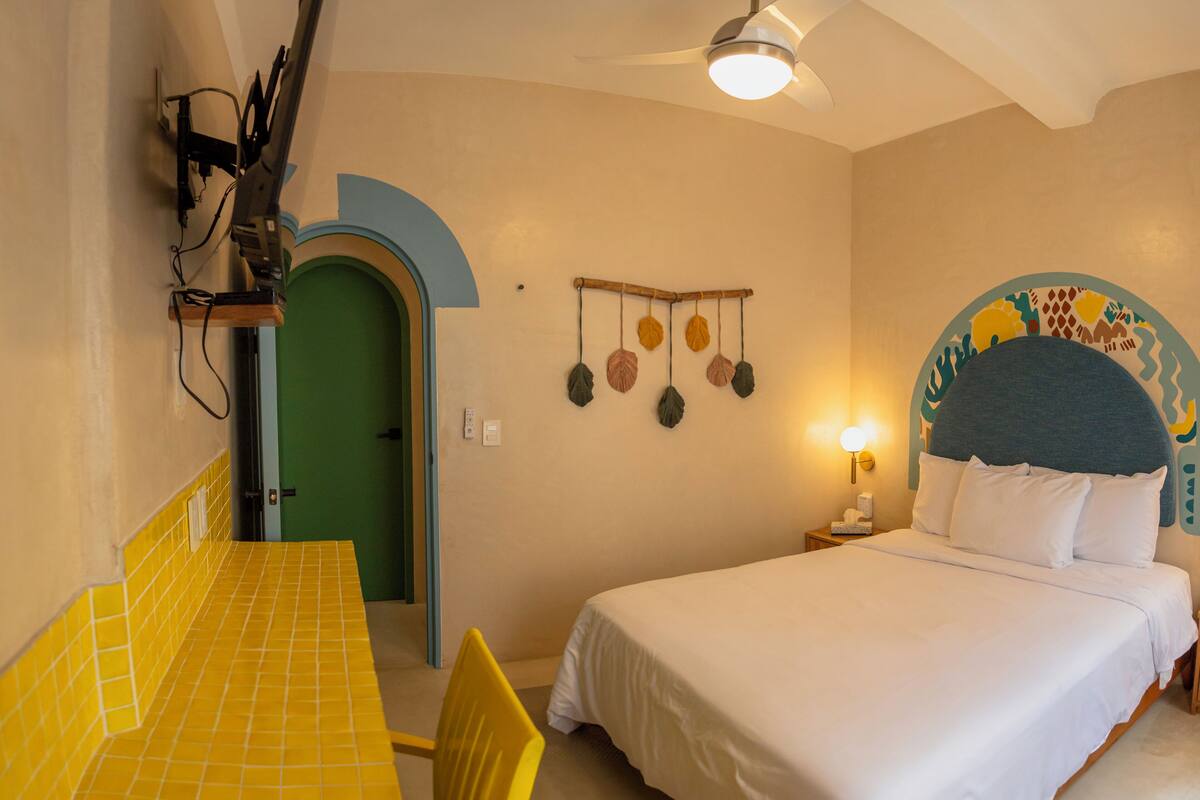 Quarto de hotel com cama, escrivaninha, cadeira, porta verde, luminária na parede e ventoinha no teto.