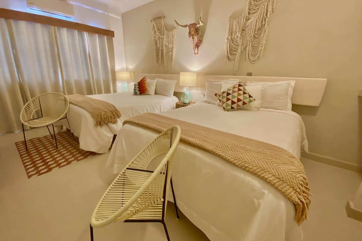 Quarto de hotel com duas camas, uma cadeira branca, um painel de macramé na parede e um tapete estampado.
