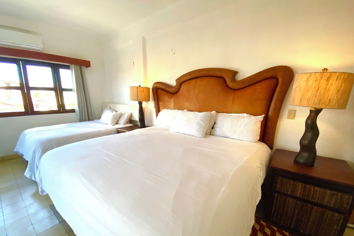 Quarto de hotel com duas camas, cabeceira de madeira, luminárias de cabeceira e piso xadrez.