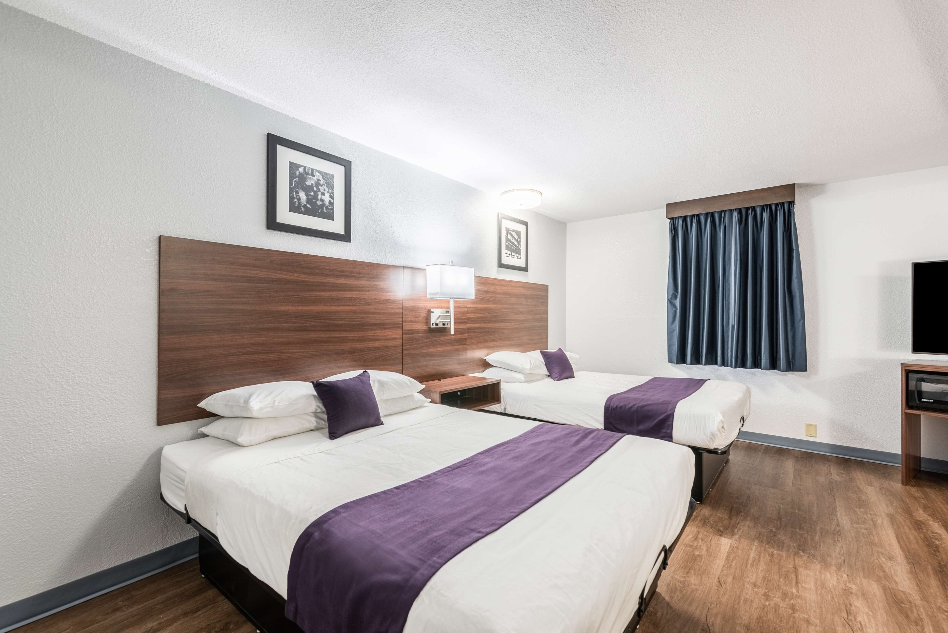Quarto de hotel com duas camas, uma cabeceira de madeira, uma mesa de cabeceira com abajur, uma televisão e uma janela com cortinas.