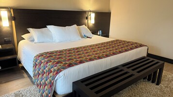 Cama bem arrumada com manta colorida de padrão geométrico, cabeceira escura e abajures laterais.