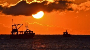 Um pôr do sol sobre o oceano, com duas silhuetas de barcos no céu.