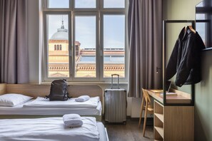 Quarto de hotel com duas camas, uma escrivaninha, uma cadeira, uma mala, uma mochila e vista para um edifício pela janela.