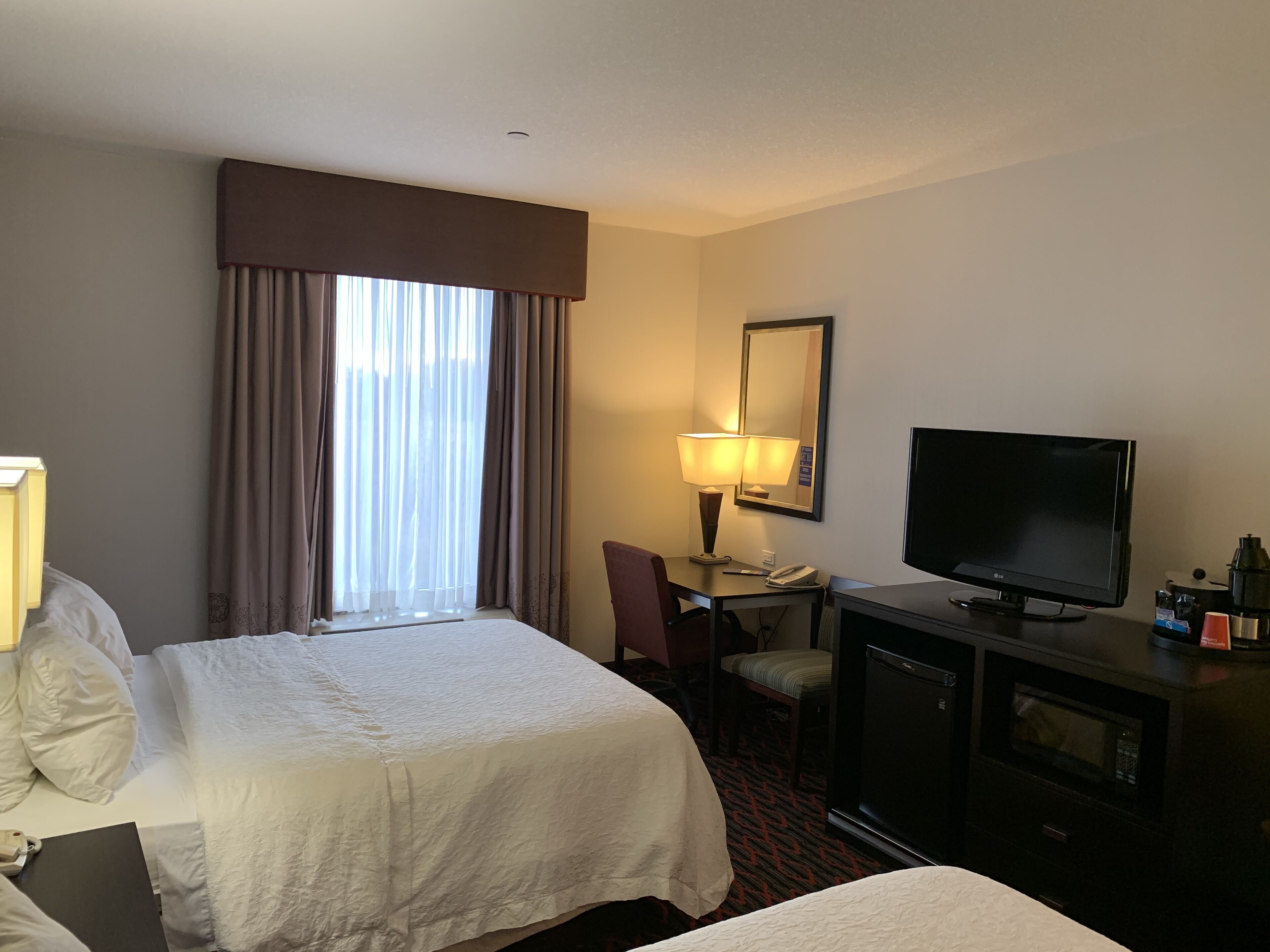 Quarto de hotel com duas camas, uma escrivaninha com luminária, televisão e minibar.
