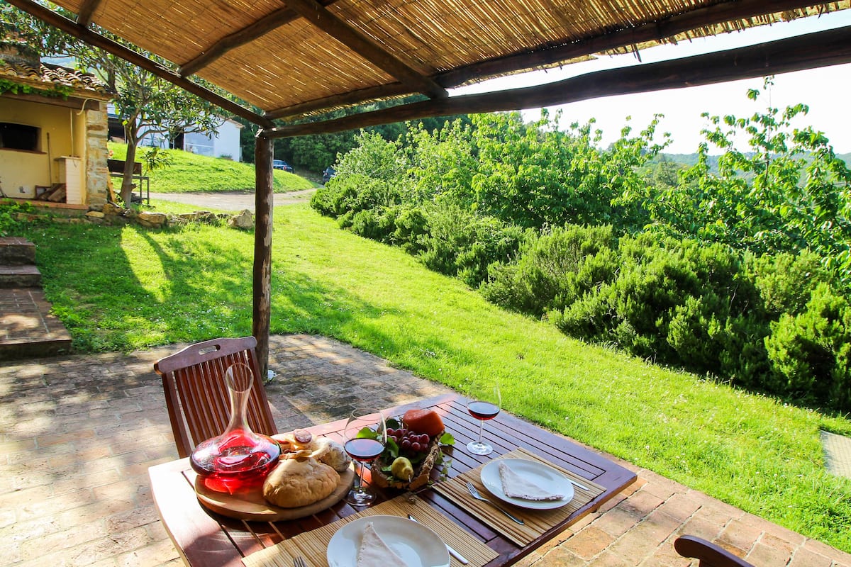 Uma área de refeições ao ar livre com uma mesa posta para dois, cercada por vegetação exuberante e vista para um vinhedo.