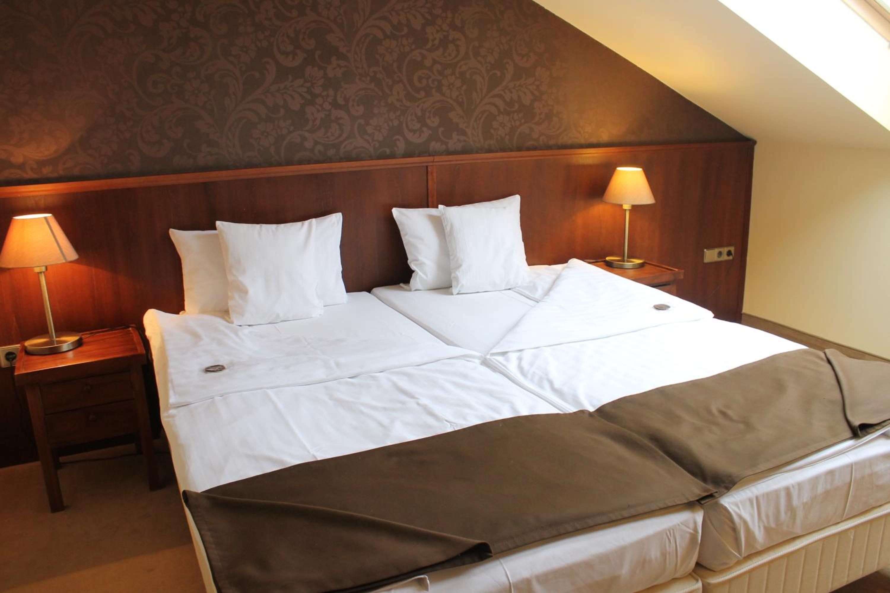 Quarto de hotel com uma cama grande, duas luminárias de cabeceira, uma cabeceira de madeira e um papel de parede estampado.