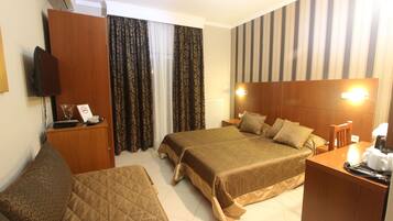 Quarto de hotel com uma cama grande, uma cama menor, uma escrivaninha com cadeira, uma televisão e uma janela com cortinas.