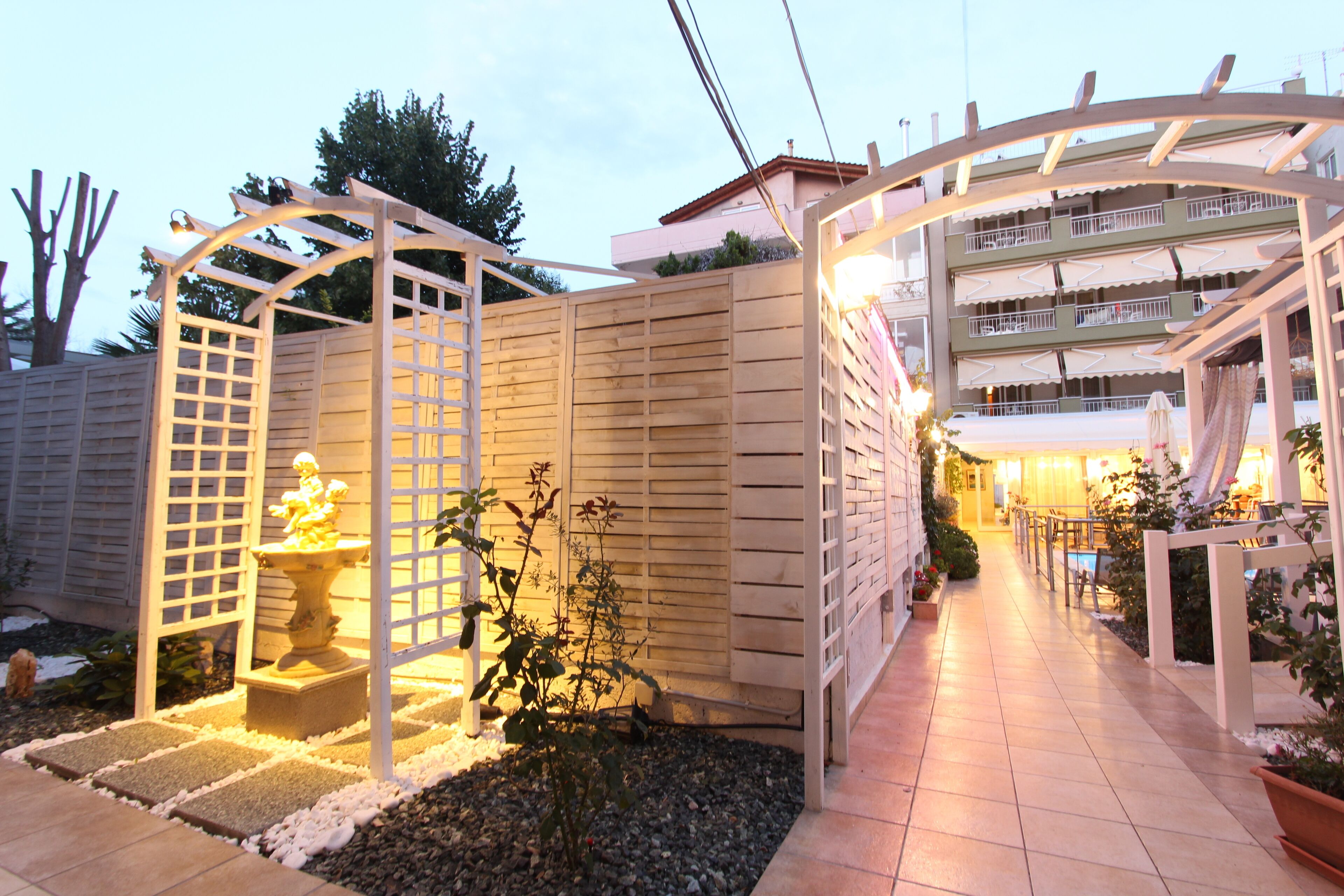 Uma área externa coberta com pérgola de madeira, uma fonte e um caminho que leva a um edifício com várias varandas.