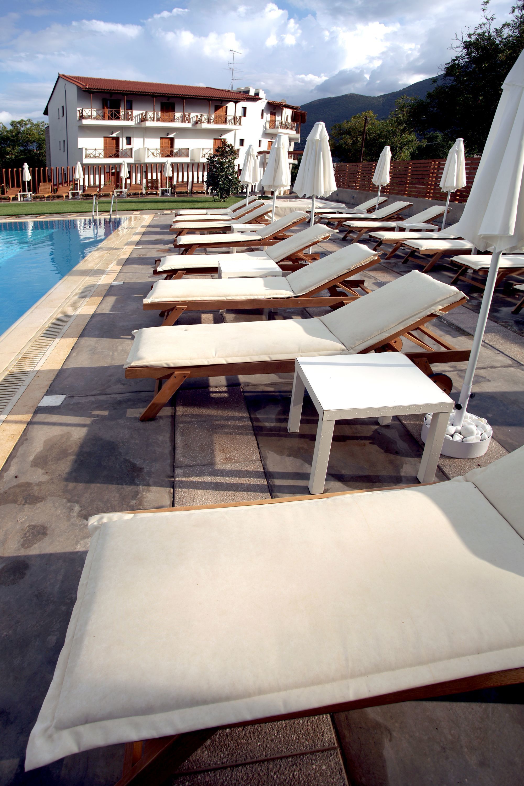 Uma área à beira da piscina com espreguiçadeiras e guarda-sóis, uma piscina e um edifício com varandas ao fundo.