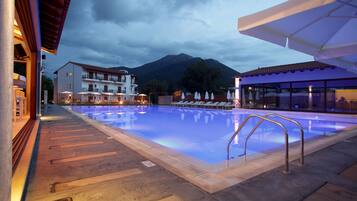 Piscina com espreguiçadeiras e guarda-sóis, cercada por edifícios e uma montanha ao fundo.