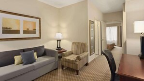 Quarto de hotel com sofá cinzento, poltrona listrada, uma mesinha com abajur, escrivaninha de madeira e um quadro na parede.