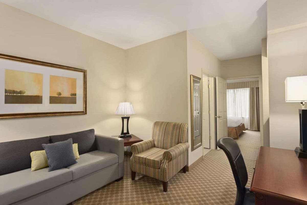 Quarto de hotel com sofá cinzento, poltrona listrada, uma mesinha com abajur, escrivaninha de madeira e um quadro na parede.