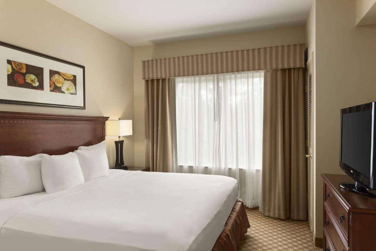 Quarto de hotel com uma cama grande, televisão de tela plana, uma obra de arte floral emoldurada e uma janela com cortinas.
