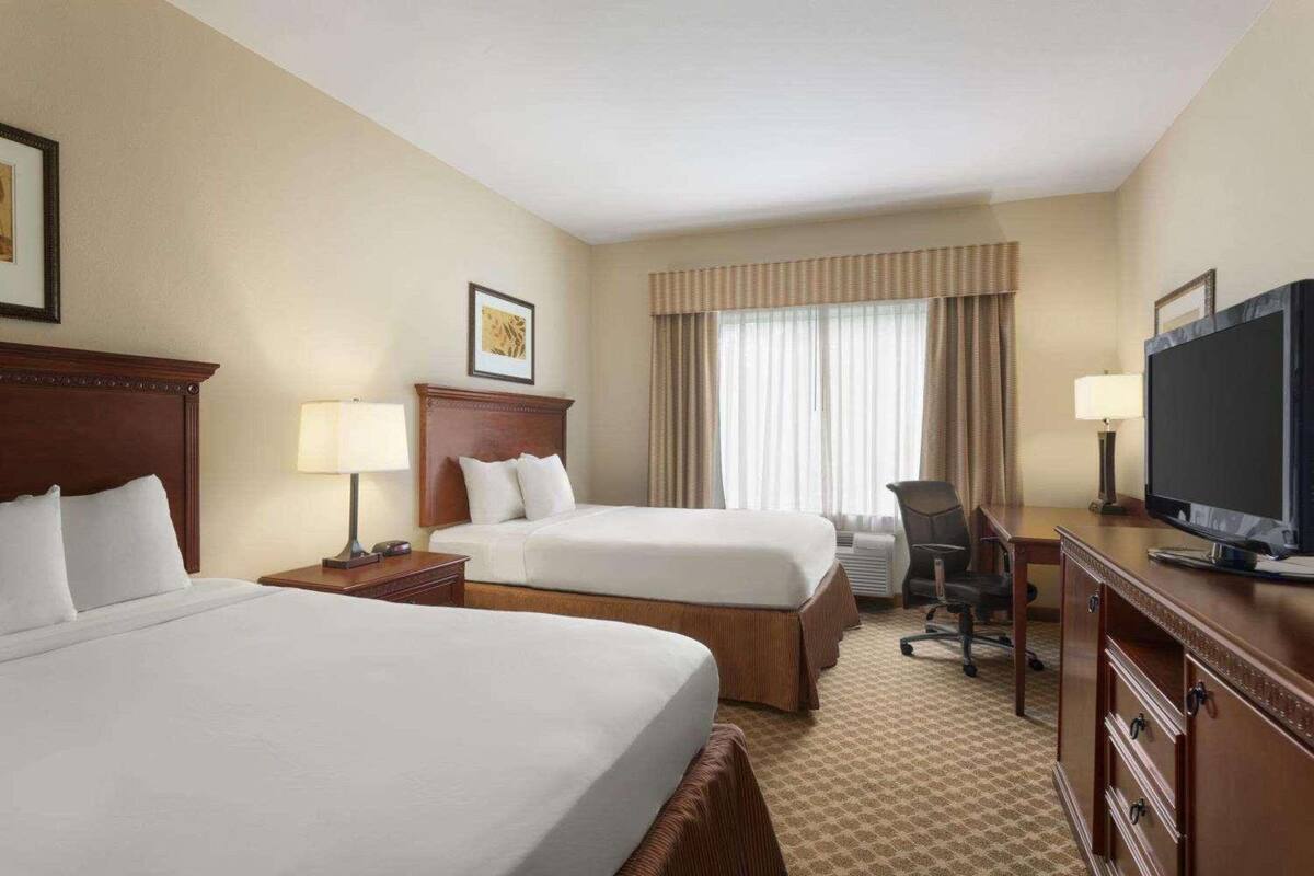 Quarto de hotel com duas camas, uma escrivaninha com televisão, uma cadeira e um abajur.