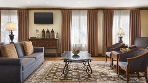 Uma sala de estar com sofá, poltronas, mesa de centro de vidro, TV na parede e um aparador com objetos decorativos.