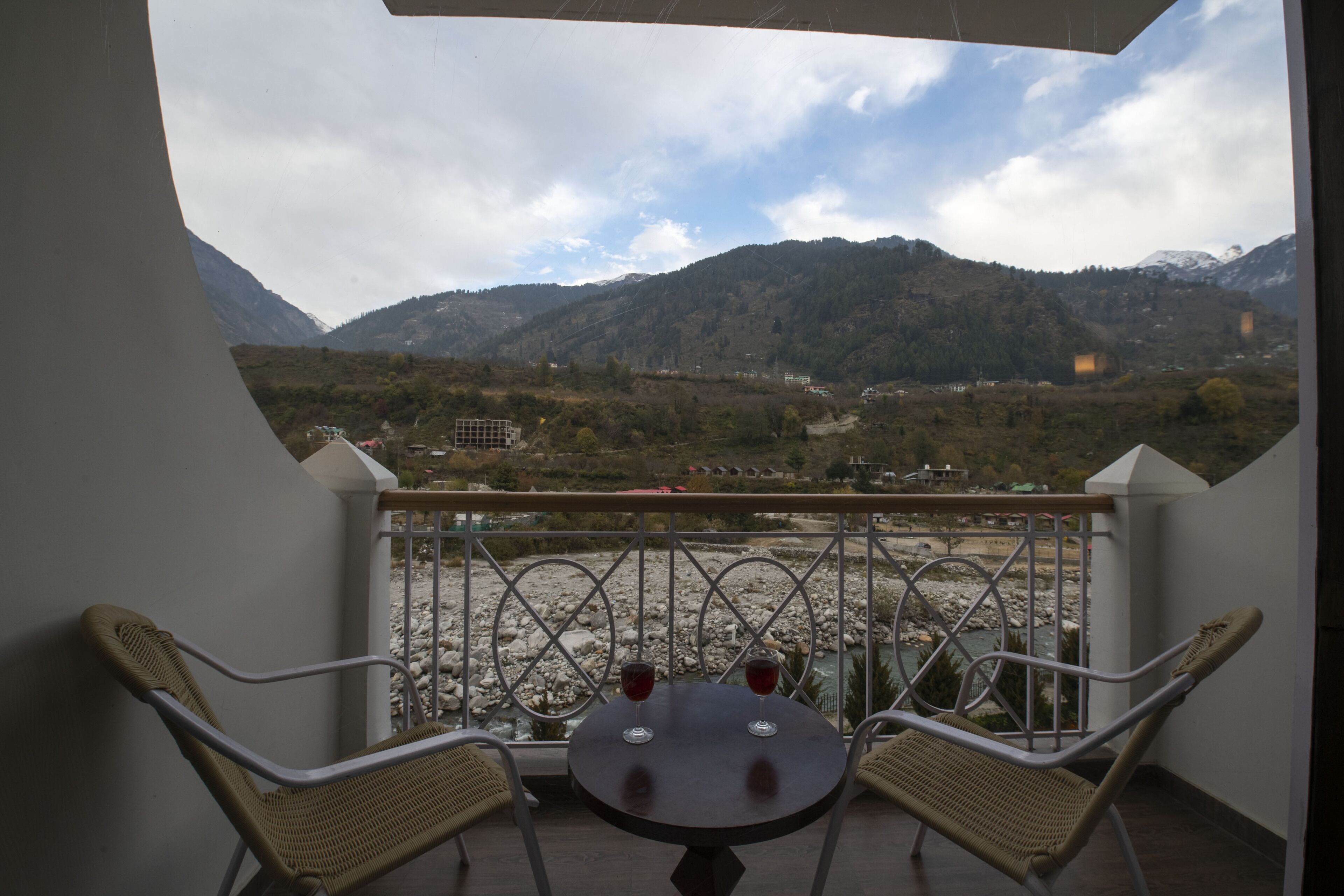Varanda com duas cadeiras de vime e uma pequena mesa, com vista para uma paisagem montanhosa com rio e edifícios.
