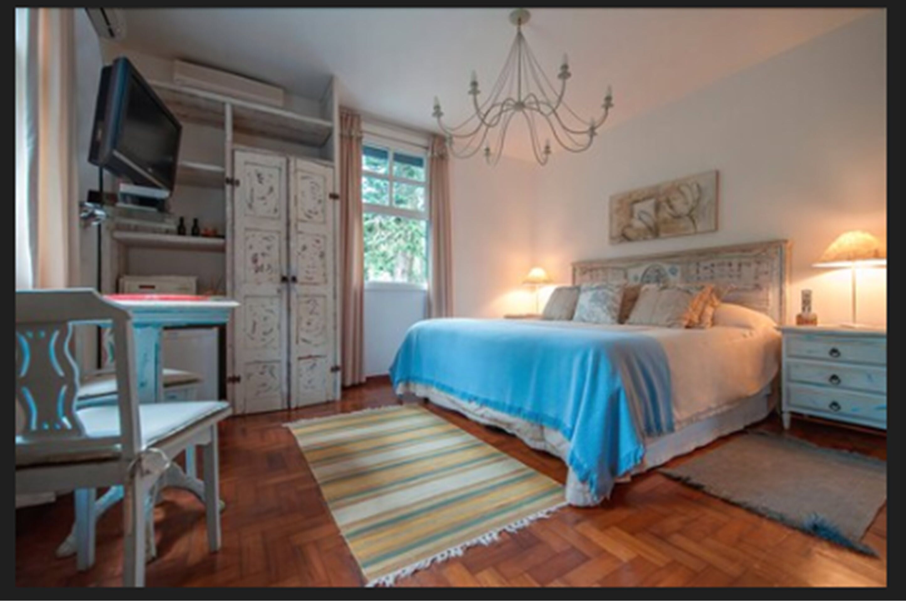 Quarto com cama, mesas de cabeceira, escrivaninha, cadeira, televisão e lustre.