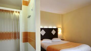 Quarto de hotel com cama, mesa de cabeceira, luminária na parede e área de chuveiro com porta de vidro.