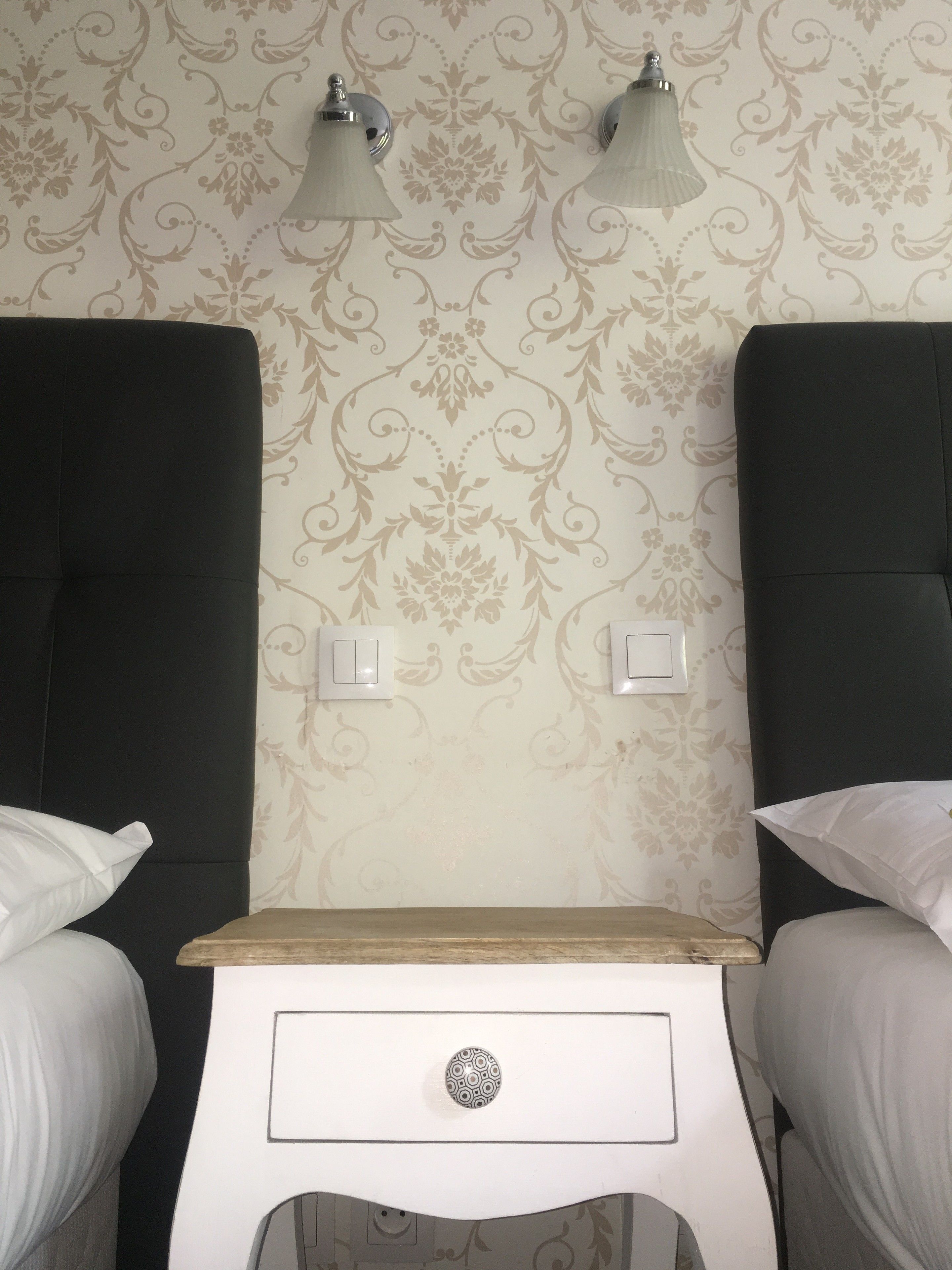 Quarto de hotel com duas camas, uma mesinha de cabeceira pequena e duas luminárias fixadas na parede.