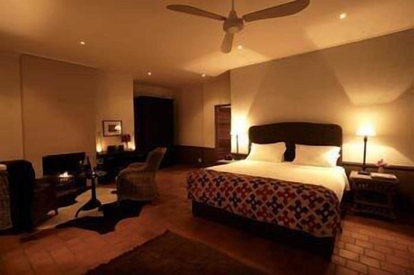 Quarto de hotel com uma cama grande, ventoinha no teto, lareira e um espaço de estar com uma poltrona e uma mesa pequena.