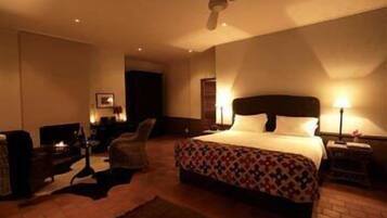 Quarto de hotel com uma cama grande, ventoinha no teto, lareira e um espaço de estar com uma poltrona e uma mesa pequena.
