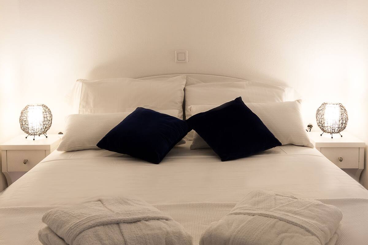 Cama bem arrumada com lençóis brancos e duas almofadas escuras, um quadro na parede e duas luminárias de cabeceira.
