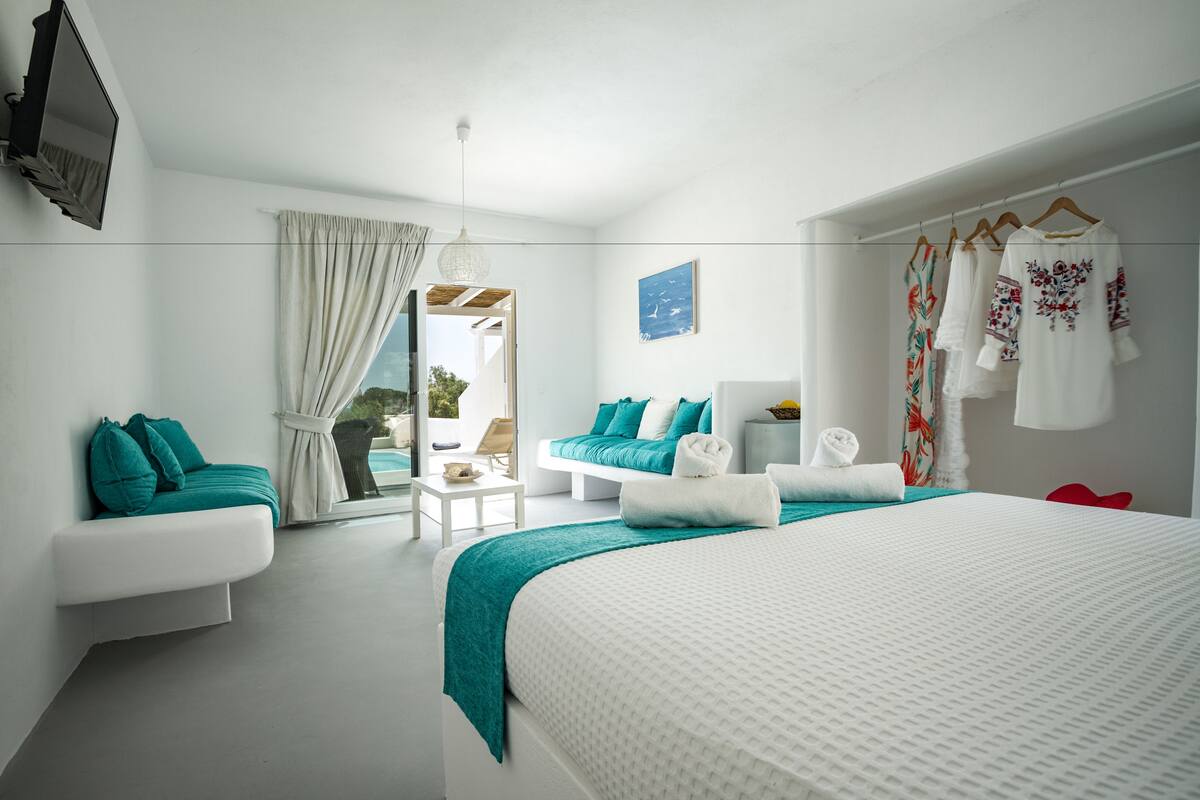 Quarto de hotel moderno com uma cama grande, um sofá e uma mesa pequena. Vê-se o exterior pela janela.