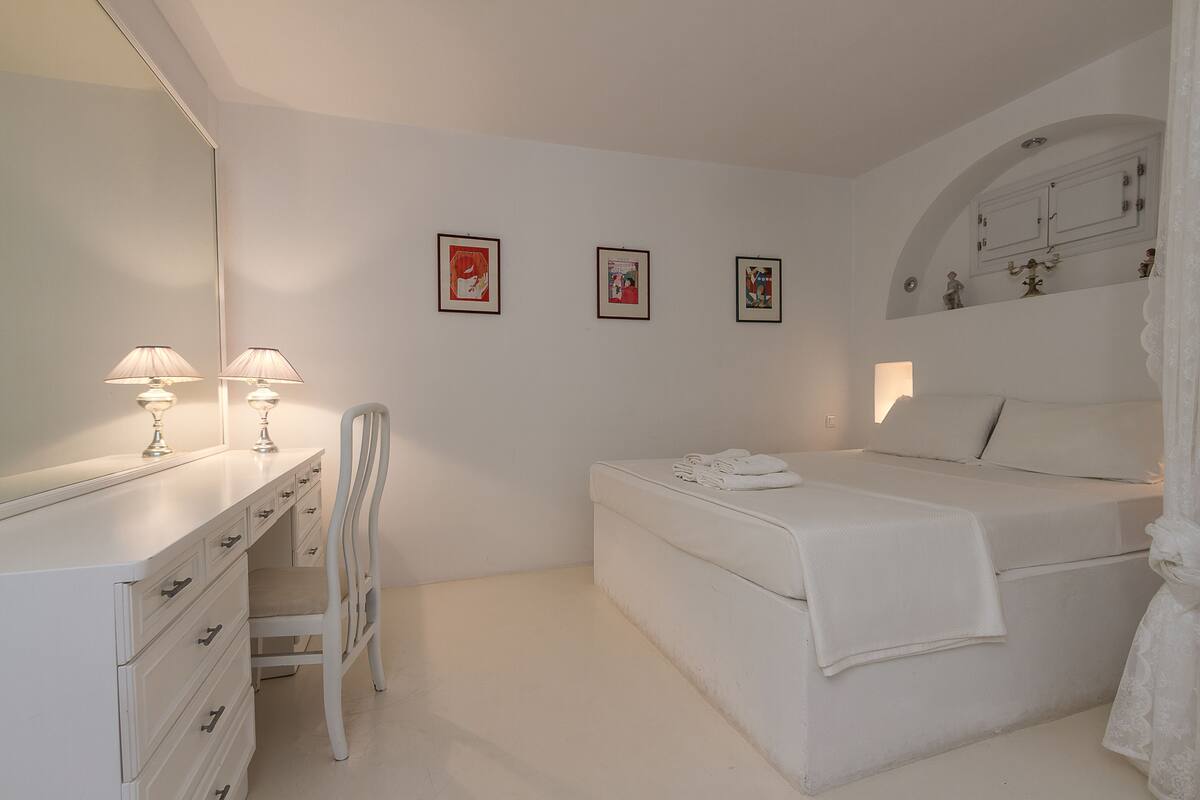 Quarto branco com cama, poltrona, cômoda e espelho. Três quadros emoldurados na parede.