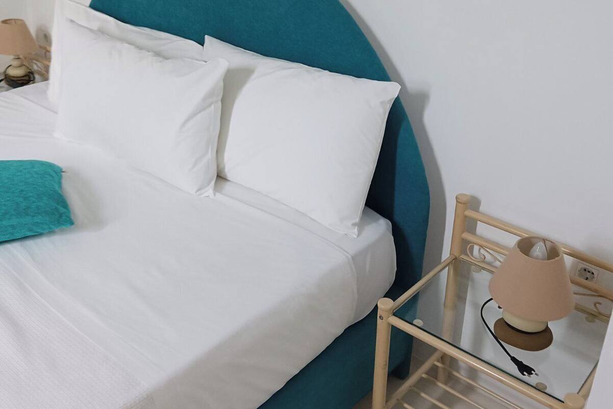 Uma cama com cabeceira azul-turquesa, roupa de cama branca e uma mesa de cabeceira com abajur.