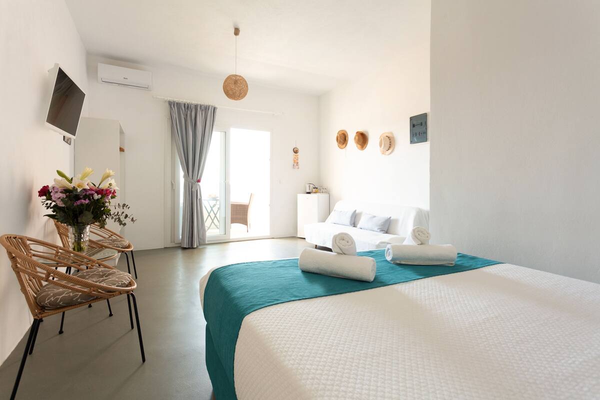 Quarto de hotel com cama, uma cadeira, mesa com flores, televisão e janela com cortinas.