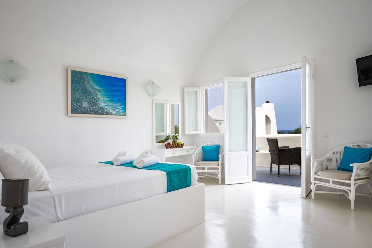 Um quarto claro e branco com uma cama, uma escrivaninha e uma cadeira. Há um quadro na parede e vista do exterior por uma porta aberta.