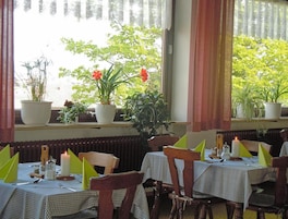 Um restaurante com mesas postas para refeição, plantas em vasos no parapeito da janela e uma vela sobre a mesa.