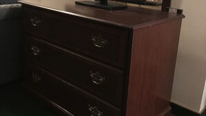 Uma cômoda de madeira com uma televisão de tela plana em cima, um espelho acima da TV e um sofá estampado ao fundo.
