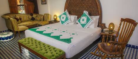Quarto bem arrumado com chão quadriculado, cama grande com roupa de cama verde e branca, um banquinho estofado verde, uma cadeira de balanço de madeira e um sofá com almofadas estampadas.