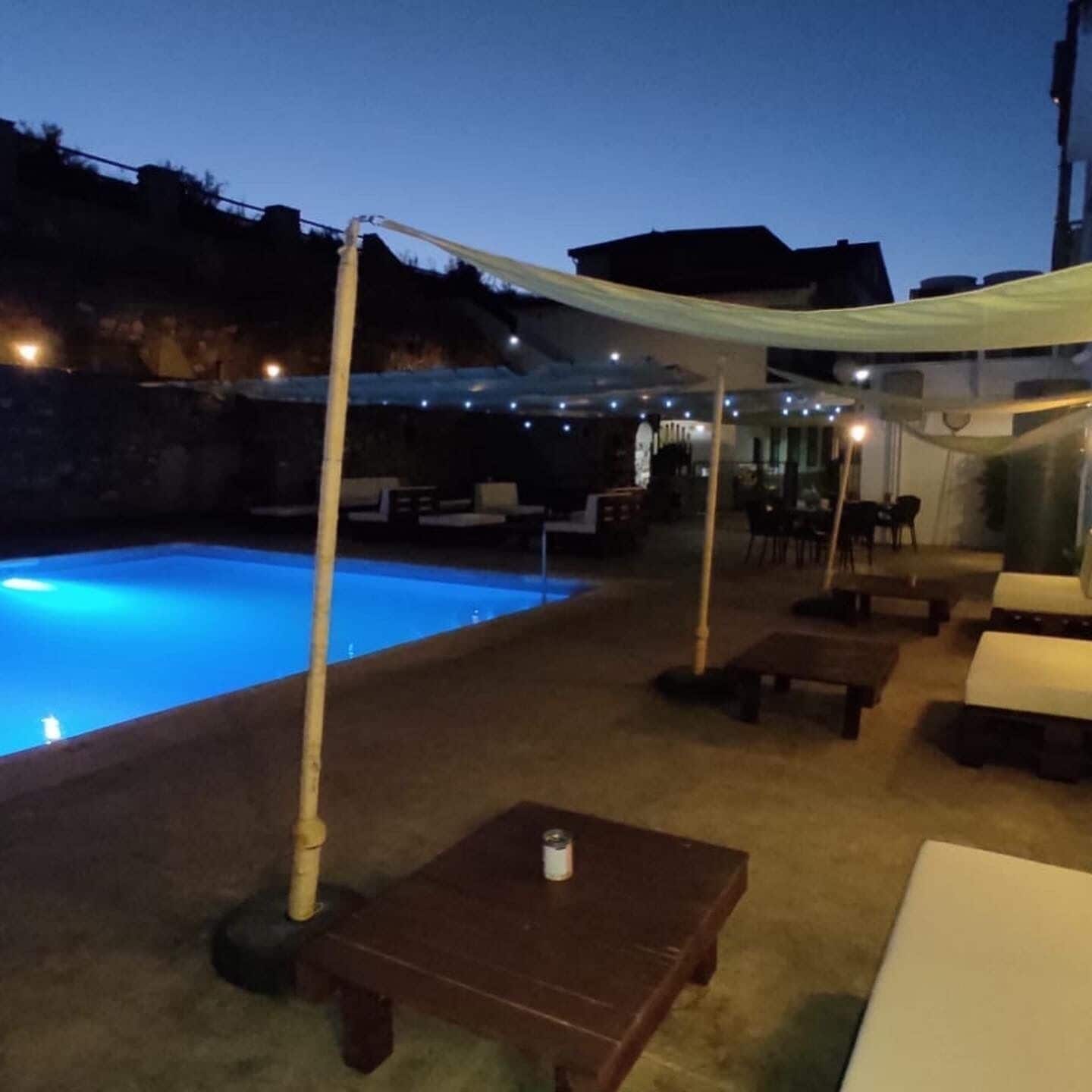 Uma área à beira da piscina com espreguiçadeiras e mesas, um espaço de estar coberto e a própria piscina.