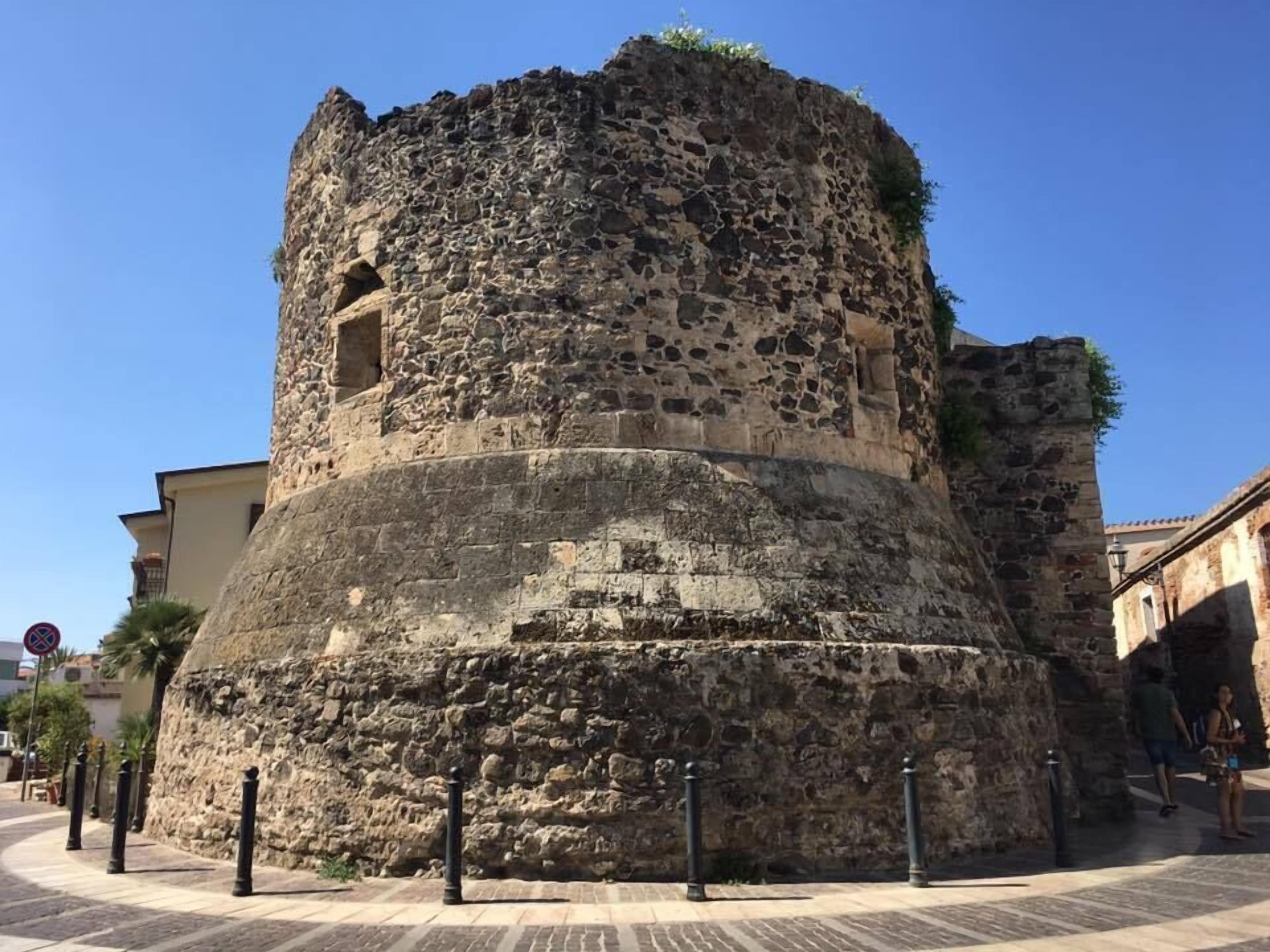 Uma torre de pedra histórica com base circular e topo plano, rodeada por uma área pavimentada com defensas.