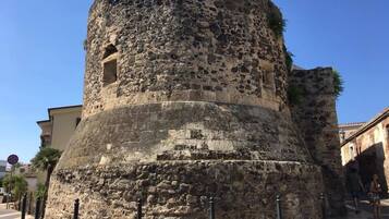 Uma torre de pedra histórica com base circular e topo plano, rodeada por uma área pavimentada com defensas.