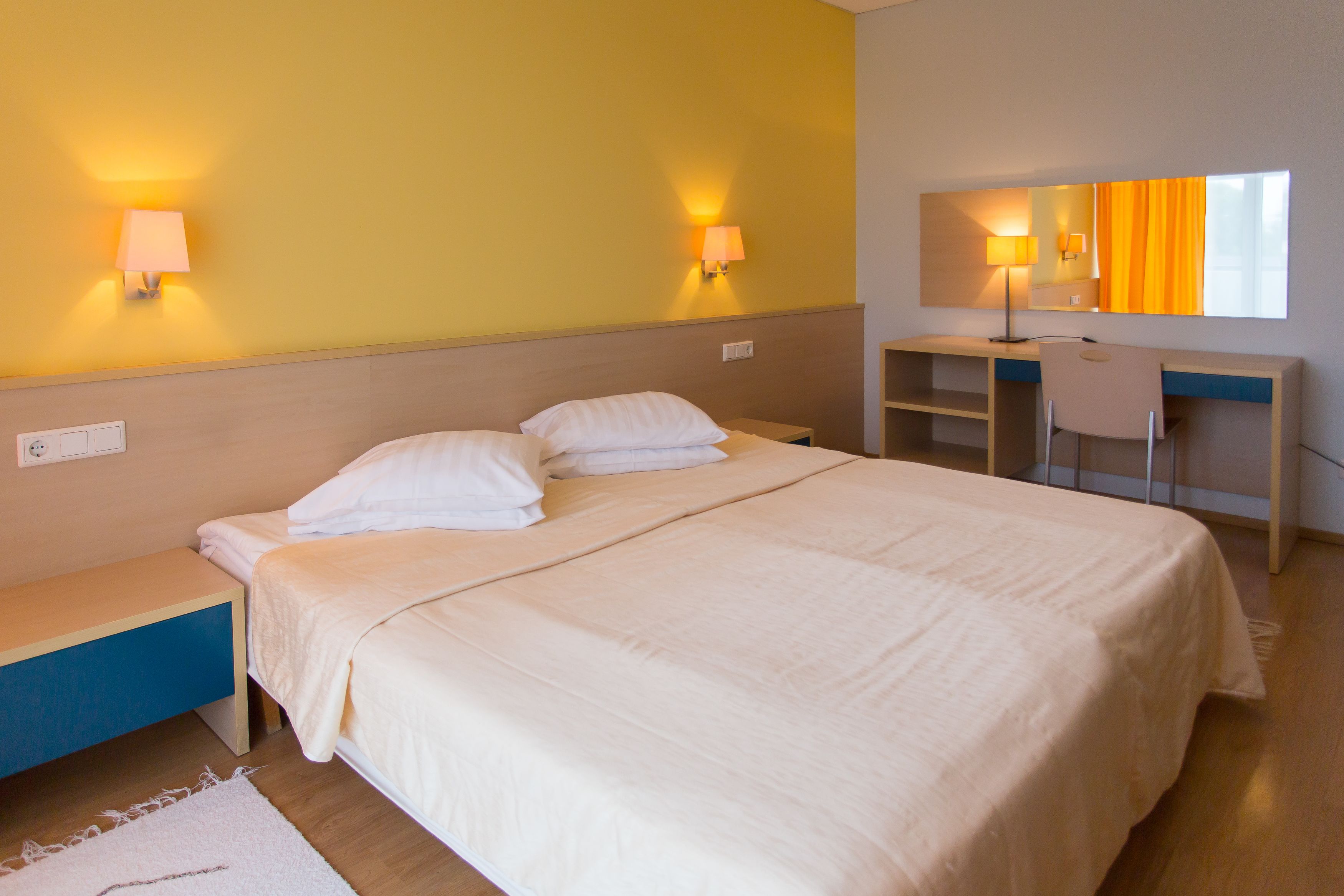 Quarto de hotel com cama, mesa de cabeceira, escrivaninha, cadeira e um espelho grande.