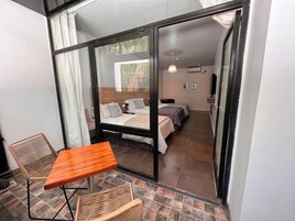 Quarto moderno com porta de vidro que dá para uma varanda, cama com roupa de cama cinza, mesa de cabeceira e luminária suspensa.