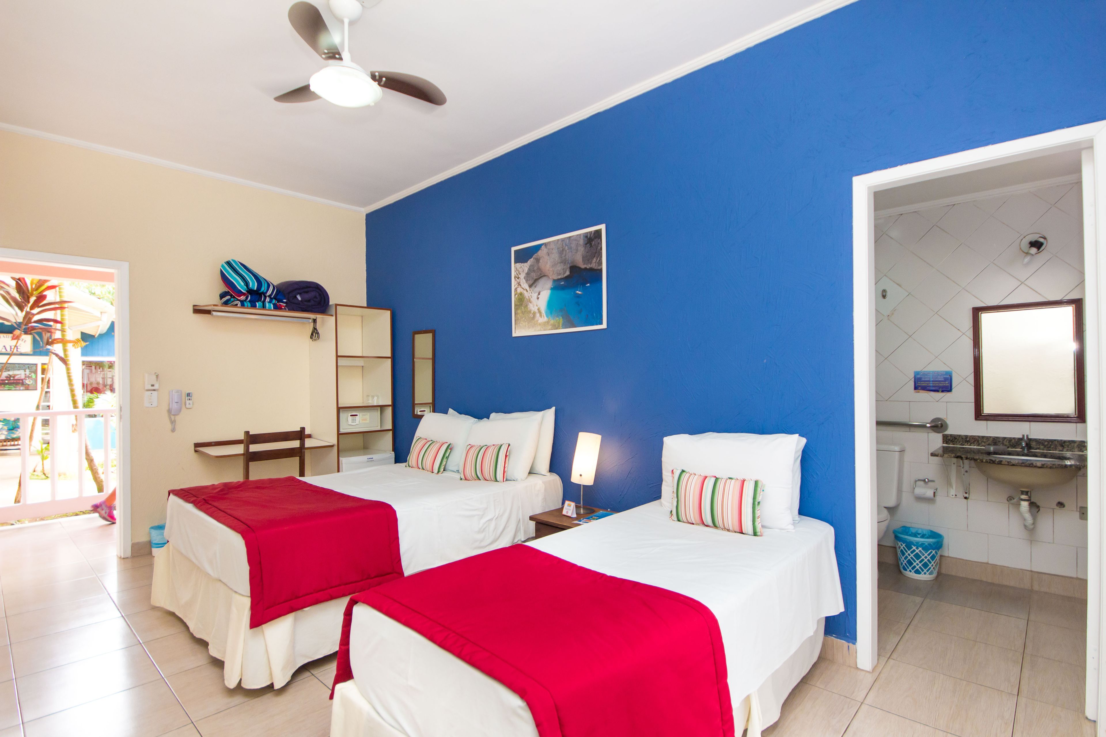 Quarto de hotel com duas camas, ventoinha no teto, banheiro com pia e espelho, e vista para o mar.