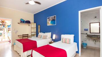 Quarto de hotel com duas camas, ventoinha no teto, banheiro com pia e espelho, e vista para o mar.