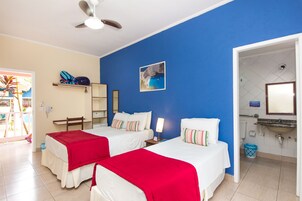 Quarto de hotel com duas camas, ventoinha no teto, banheiro com pia e espelho, e vista para o mar.