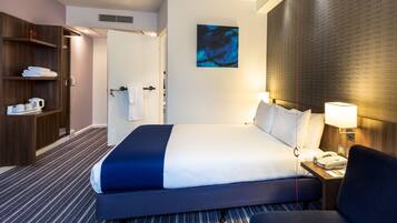 Quarto de hotel com uma cama grande, uma mesa de cabeceira com abajur, um telefone e um guarda-roupa de madeira.