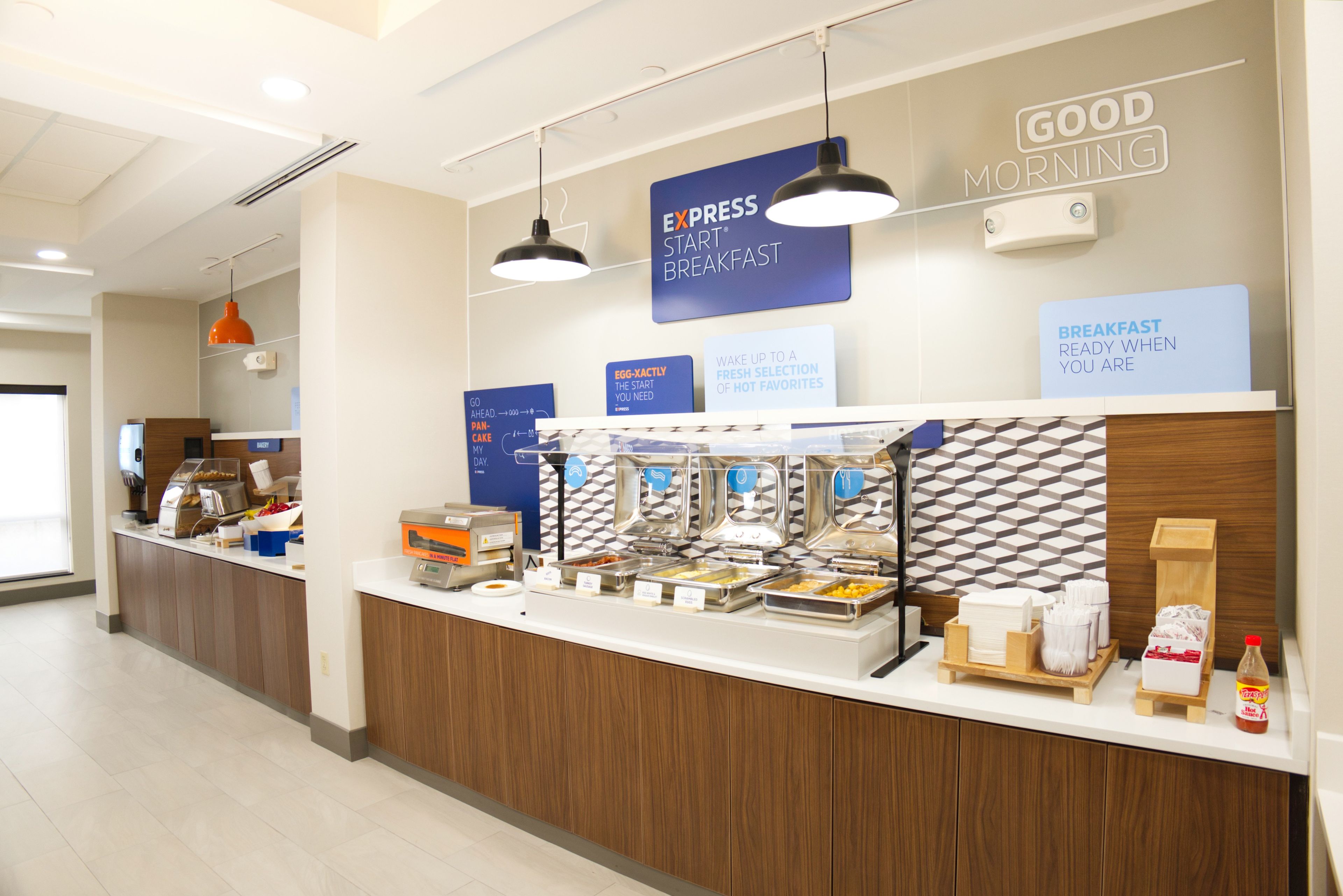 Espaço de buffet de café da manhã com diversas opções de alimentos, um letreiro com 'Bom dia' e uma placa indicando 'Café da manhã pronto quando você estiver'.