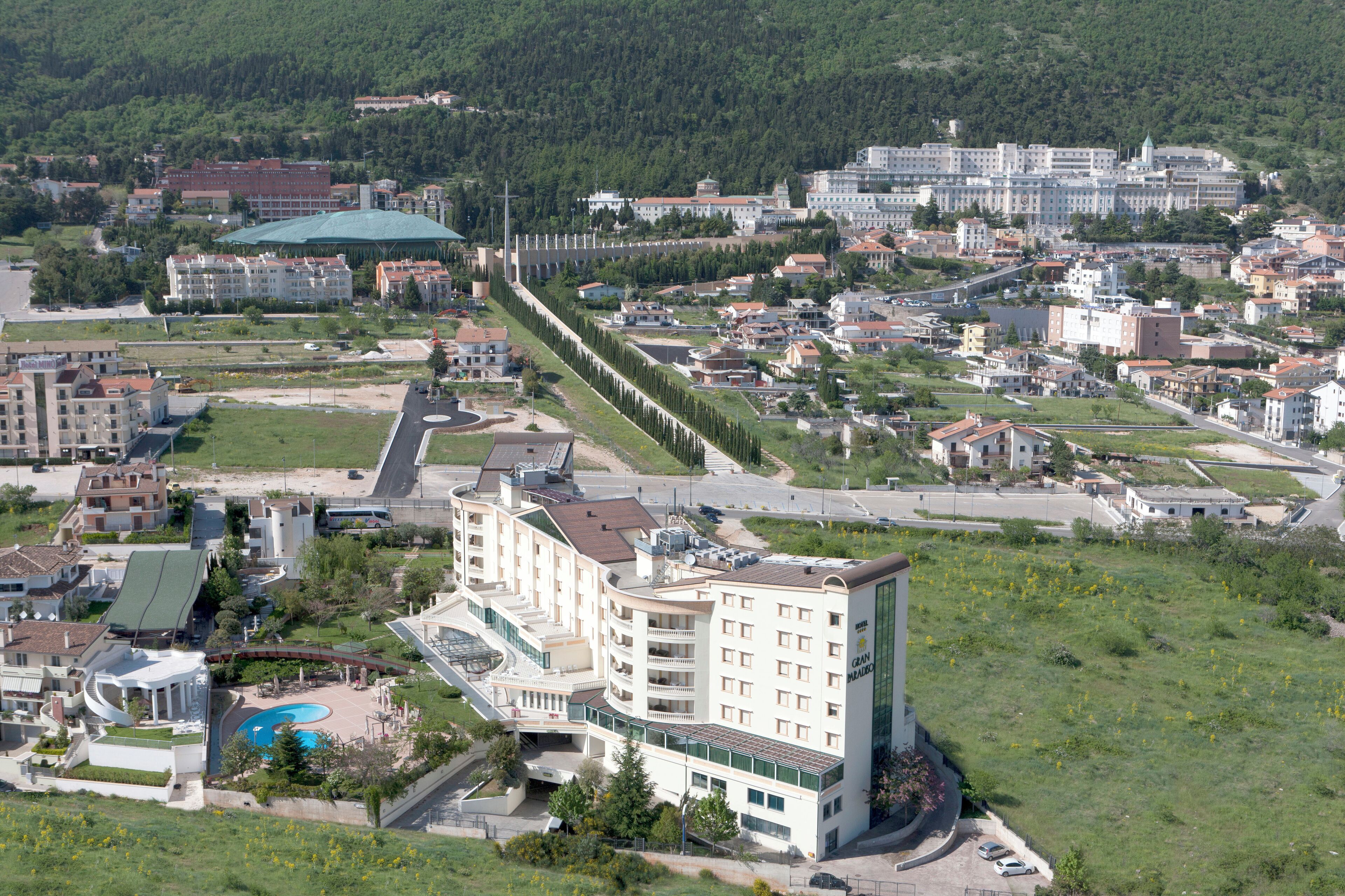 Um hotel com vários andares, piscina e vegetação exuberante ao redor, com outros edifícios nas proximidades.