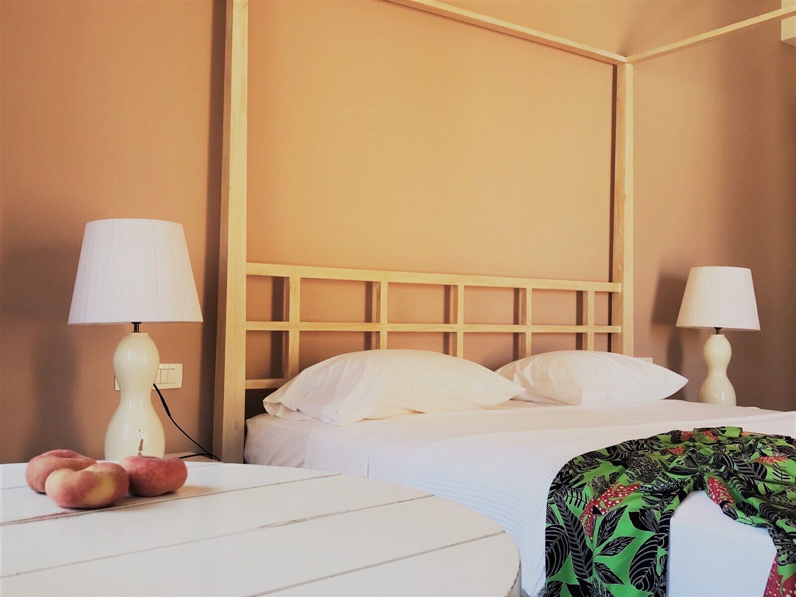 Quarto de hotel com cama, duas luminárias de cabeceira e uma mesa com frutas.